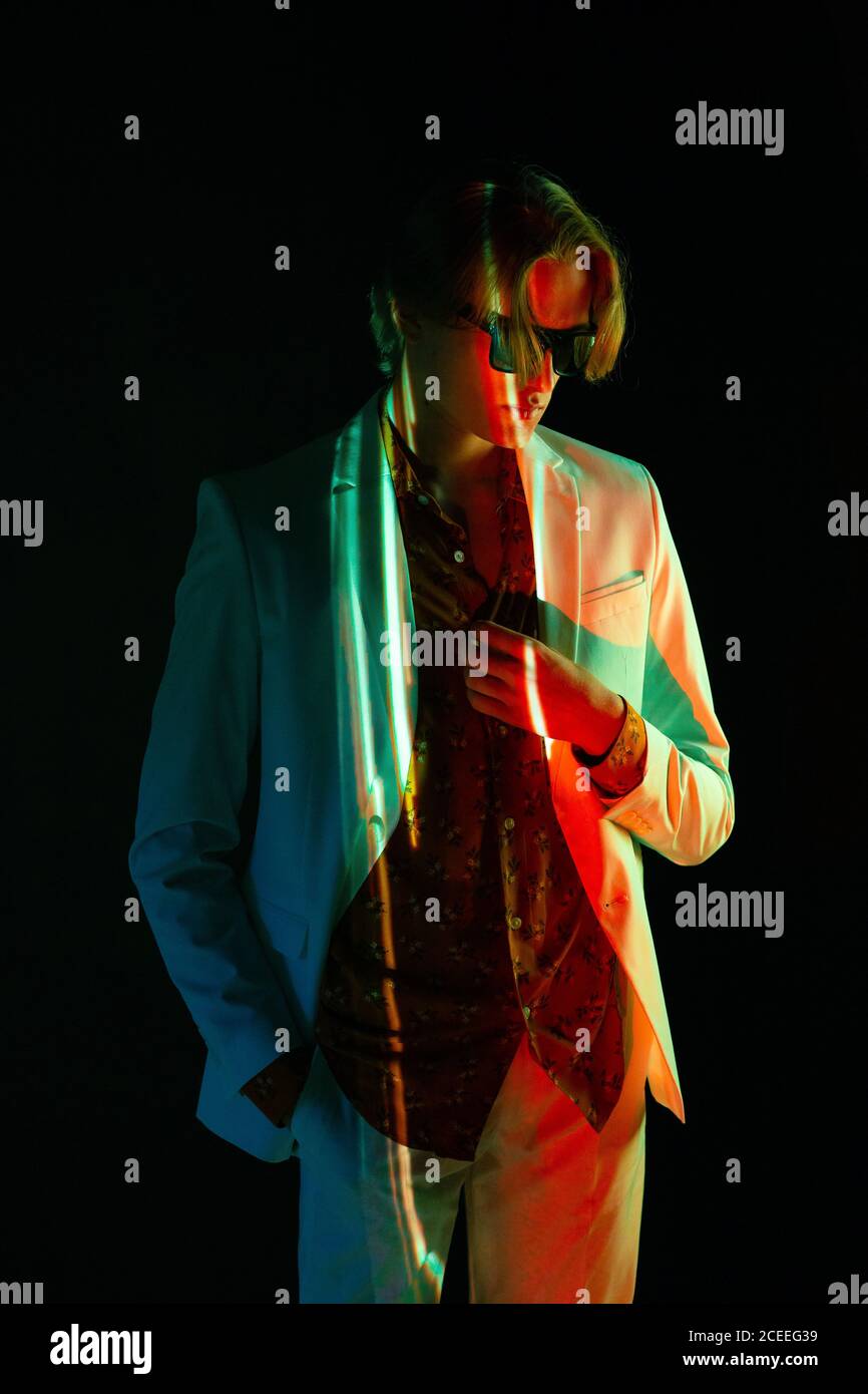 Androgynous male model in suit standing in relaxed pose under colorful illumination on black ...