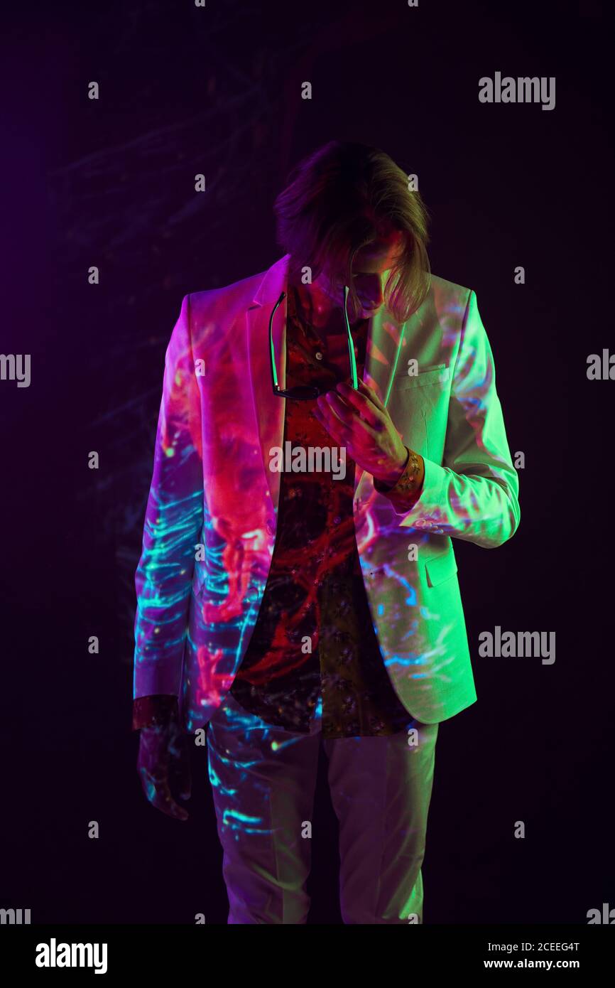 Androgynous male model in suit standing in relaxed pose under colorful illumination on black ...