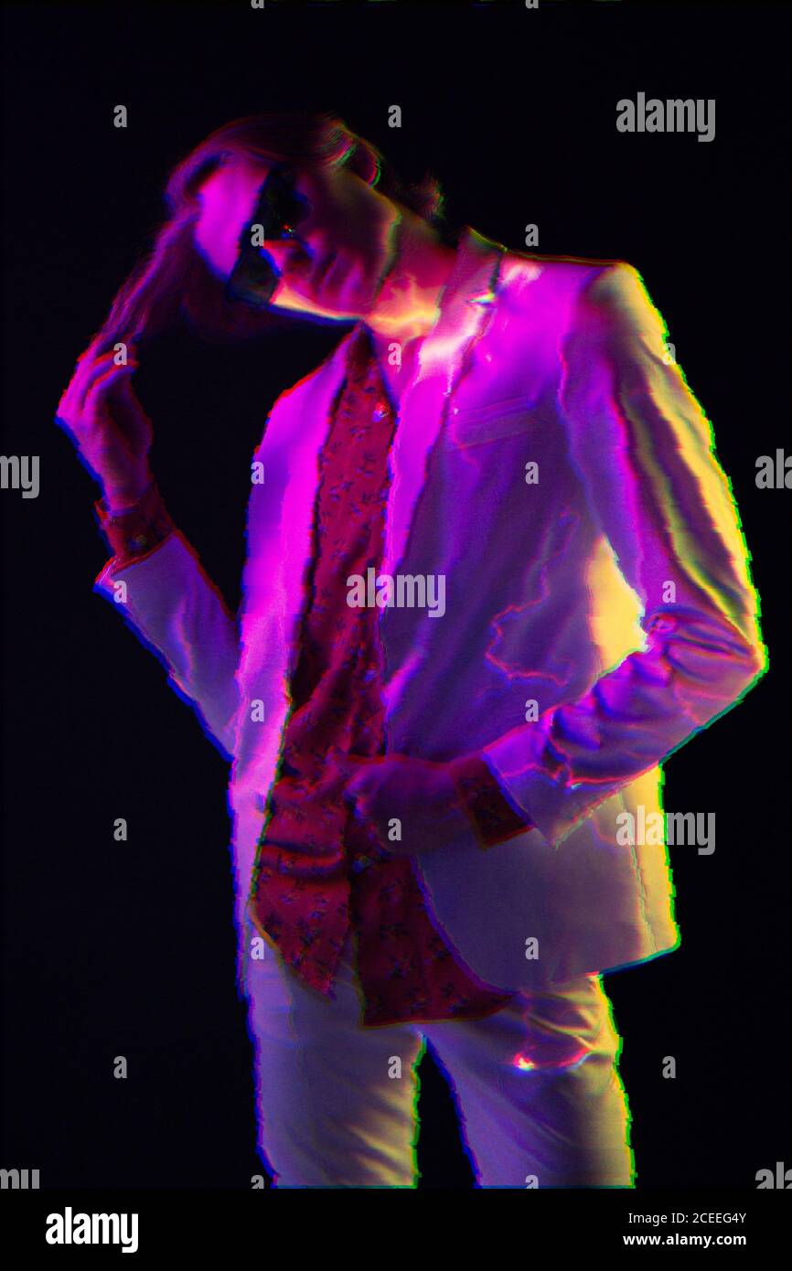 Androgynous male model in suit standing in relaxed pose under colorful illumination on black ...