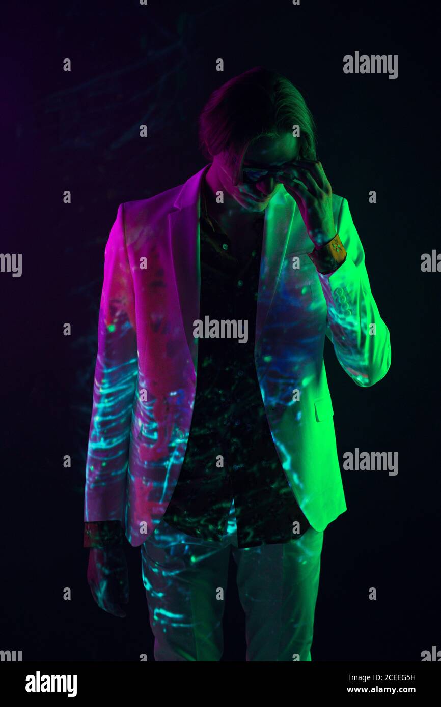 Androgynous male model in suit standing in relaxed pose under colorful illumination on black ...