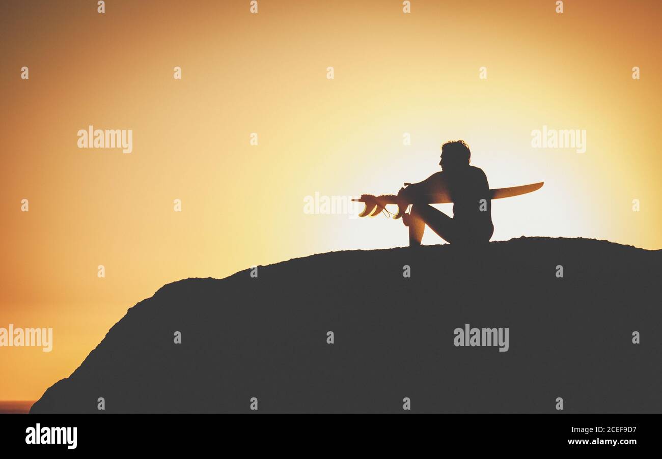 Side view of male surfer dark figure sitting on hill and holding board ...