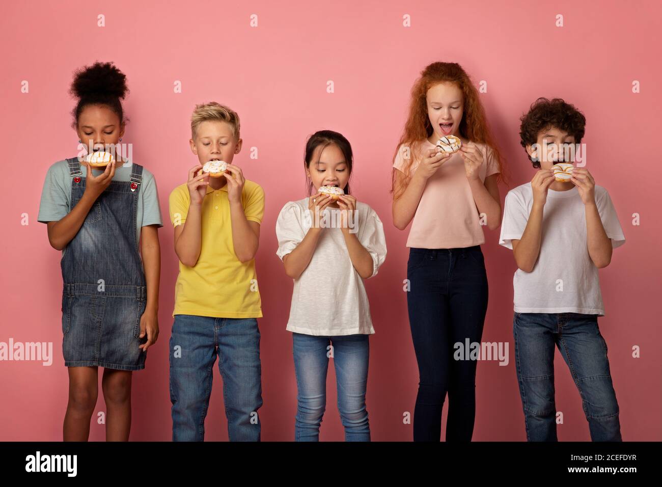 Adorable multiethnic children eating delicious sweet donuts over pink ...