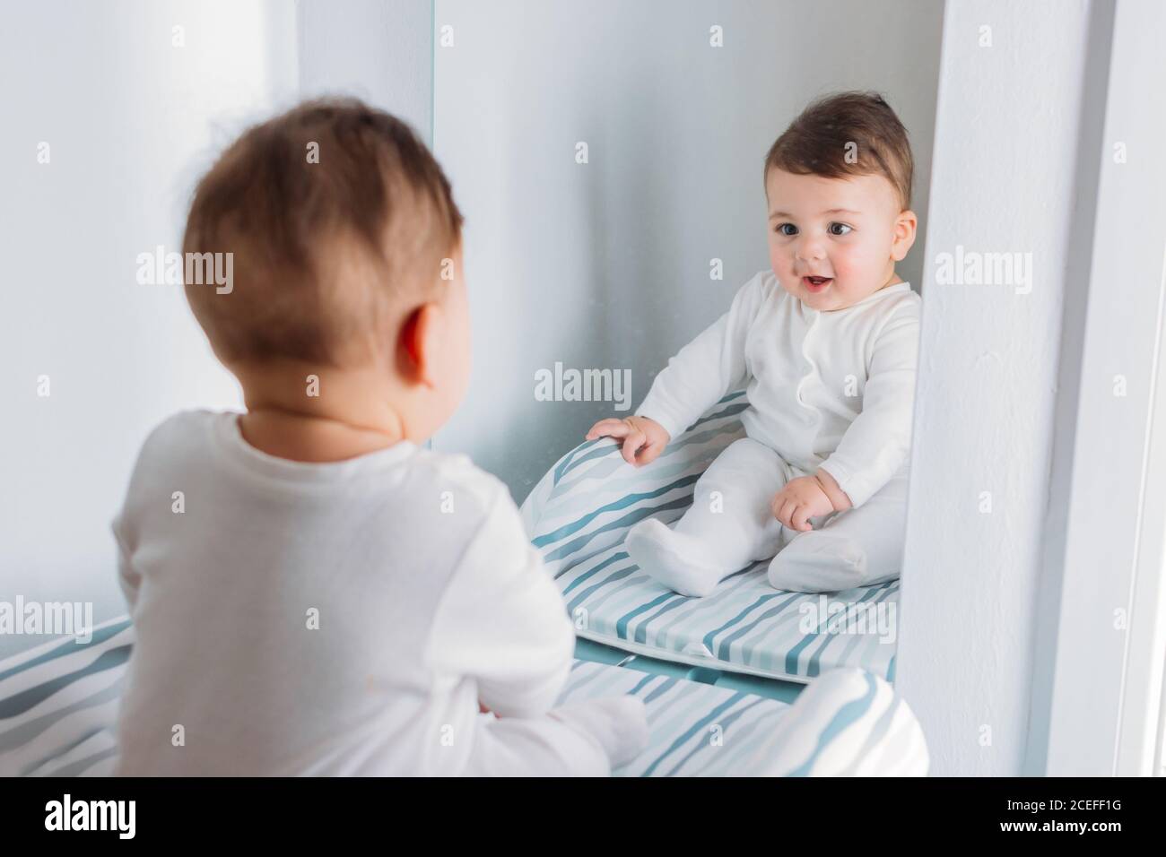 Sweet baby sitting on mat near mirror and looking at its own reflection ...
