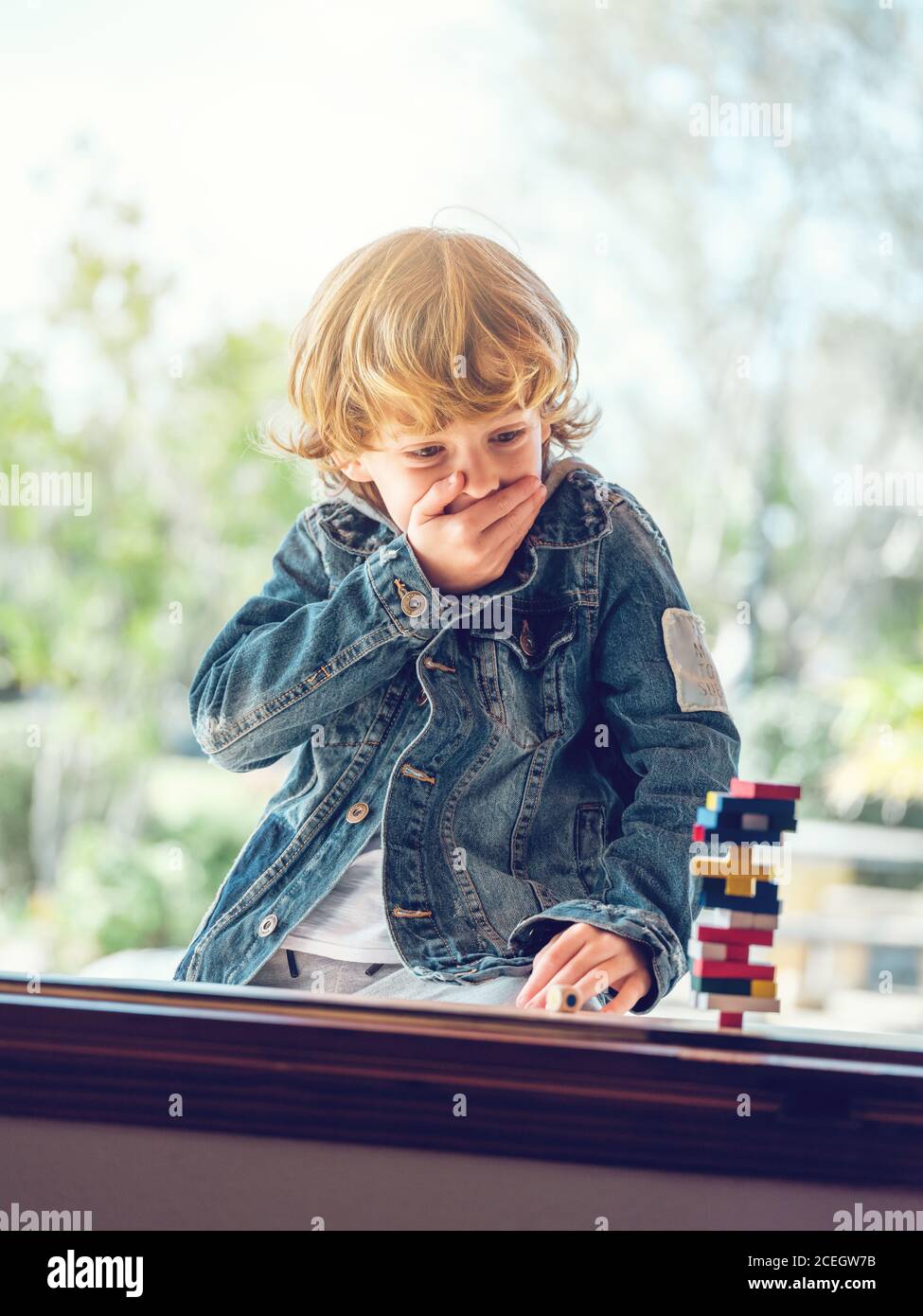 Cute surprised boy sitting at the window and playing with wooden tower ...