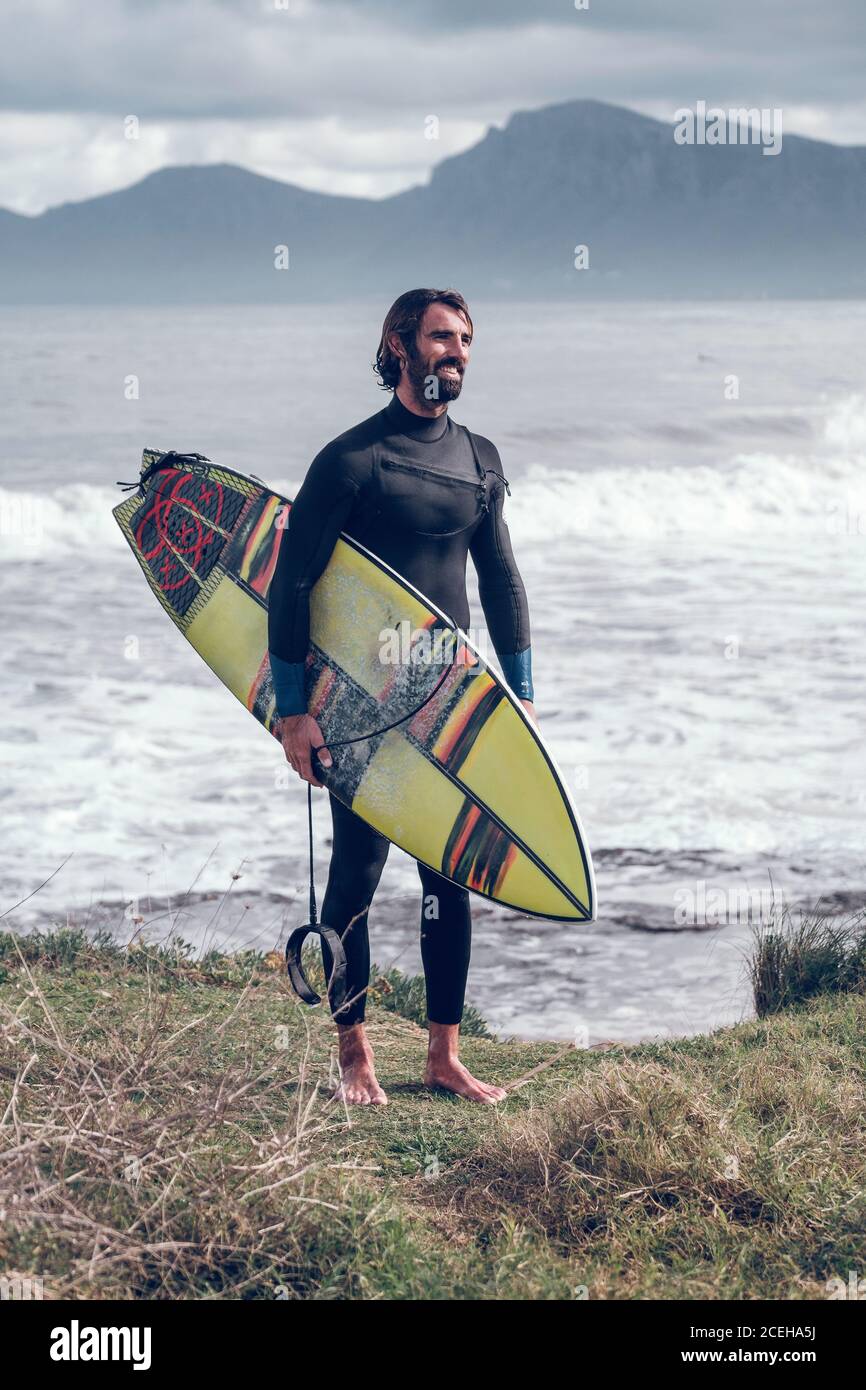 Smiling guy in diving suit standing with bright surf board on coast ...