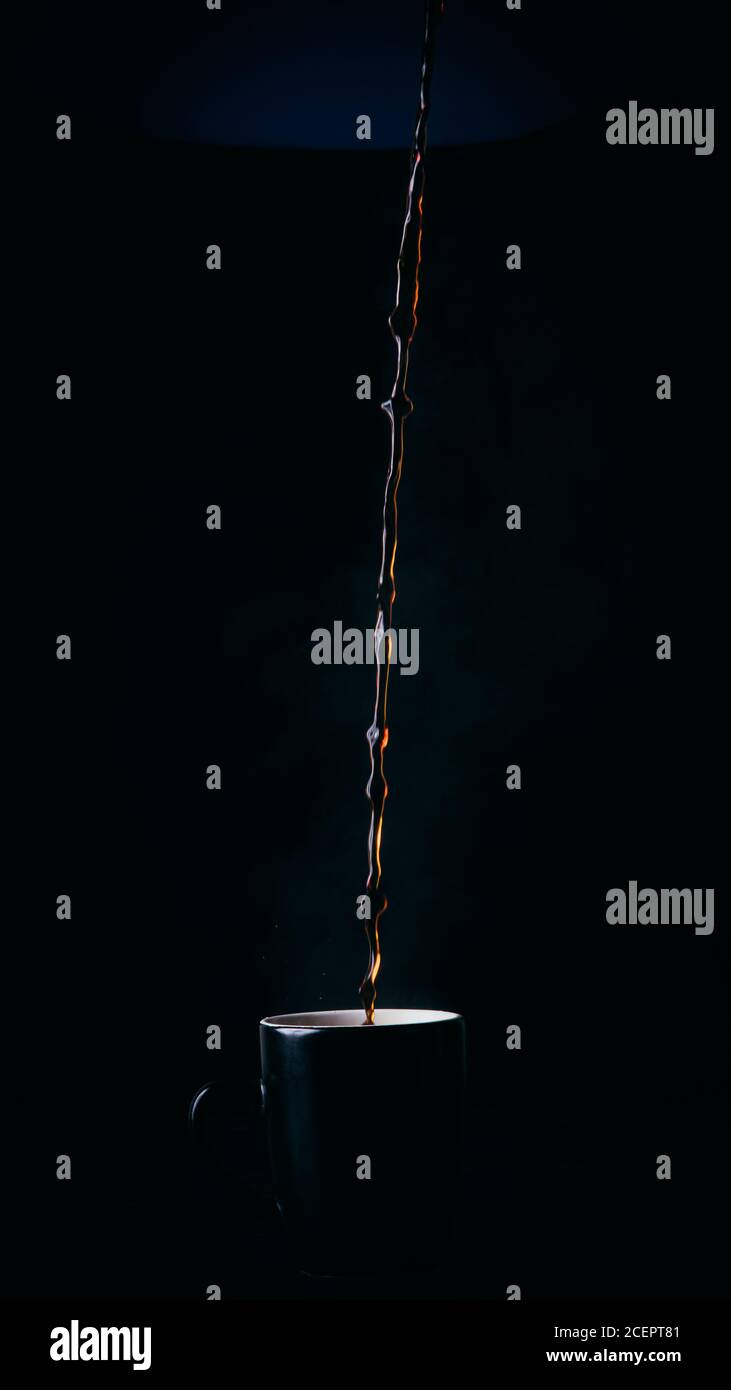 Vertical shot of coffee pouring into a black mug on a black background ...
