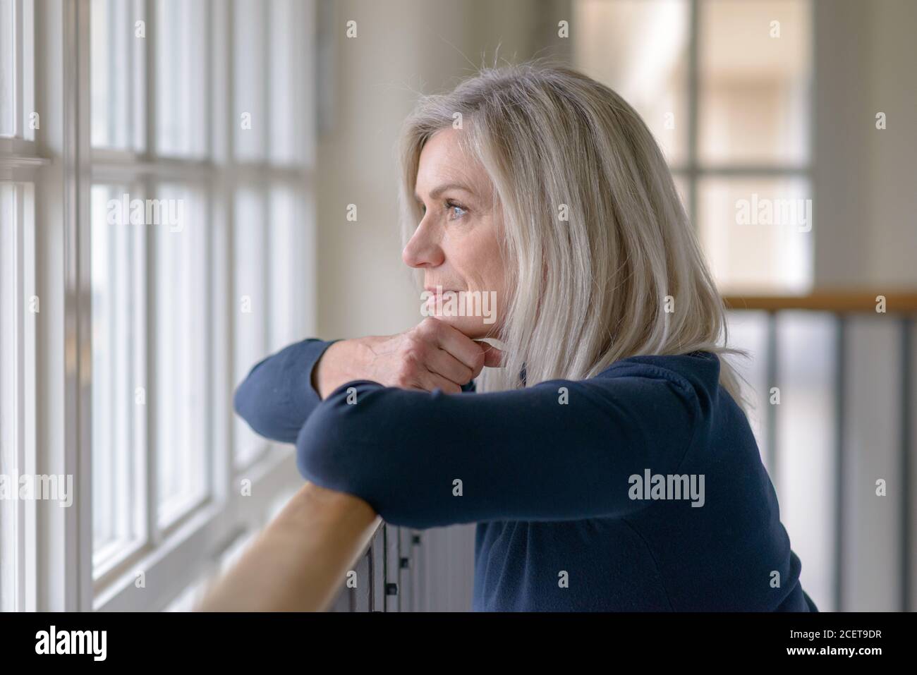 Sad blond woman watching through a window with a serious expression ...