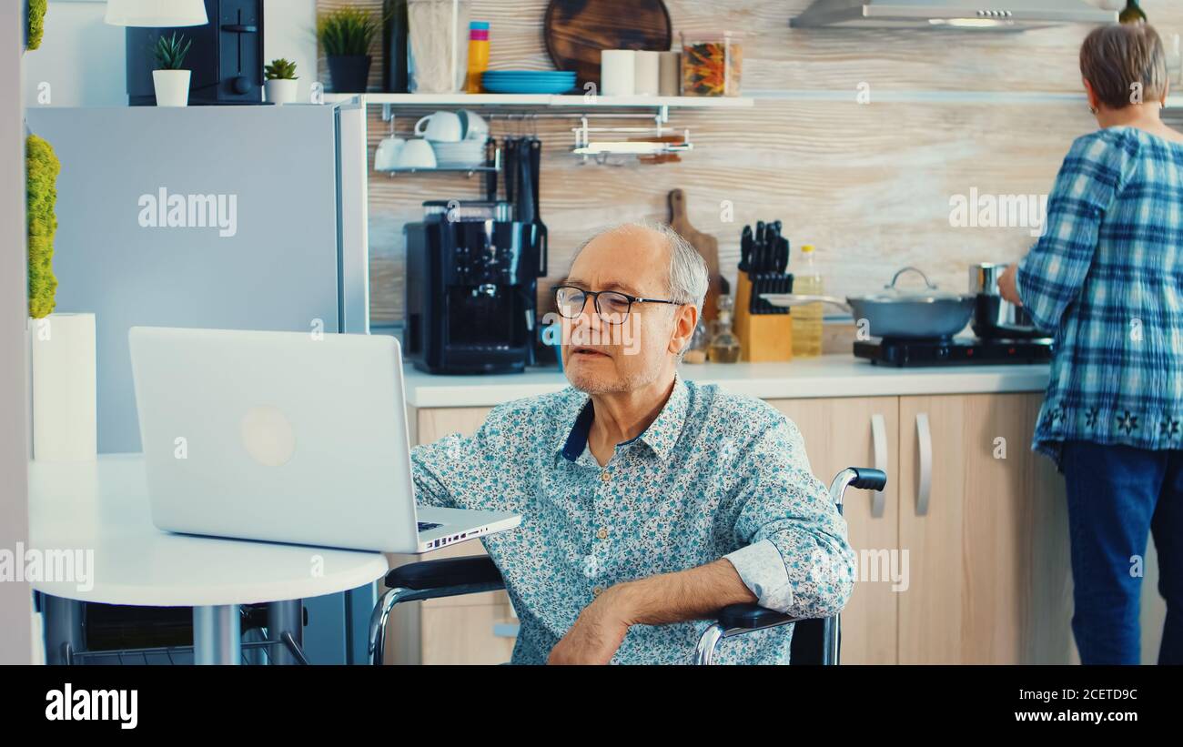 Disabled senior man in wheelchair working on laptop in kitchen while ...
