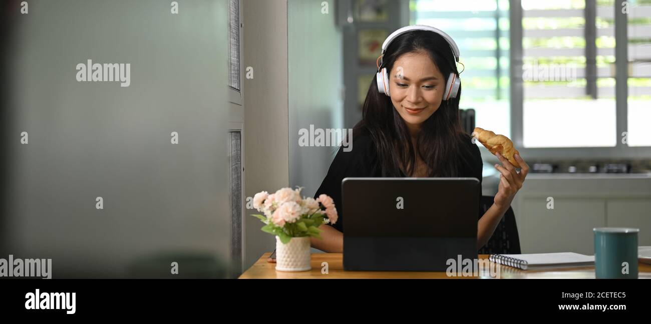 A beautiful woman is eating a croissant while using a computer tablet ...