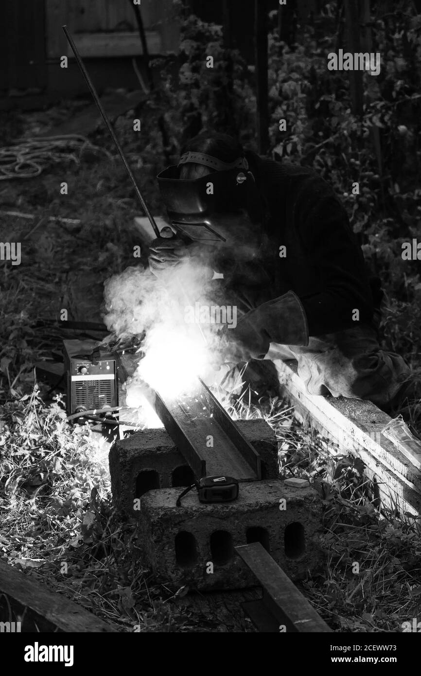 a male welder welds steel parts with electric welding in the evening at ...