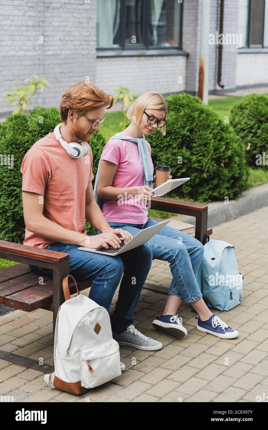 young students sitting on bench and using gadgets while online study ...