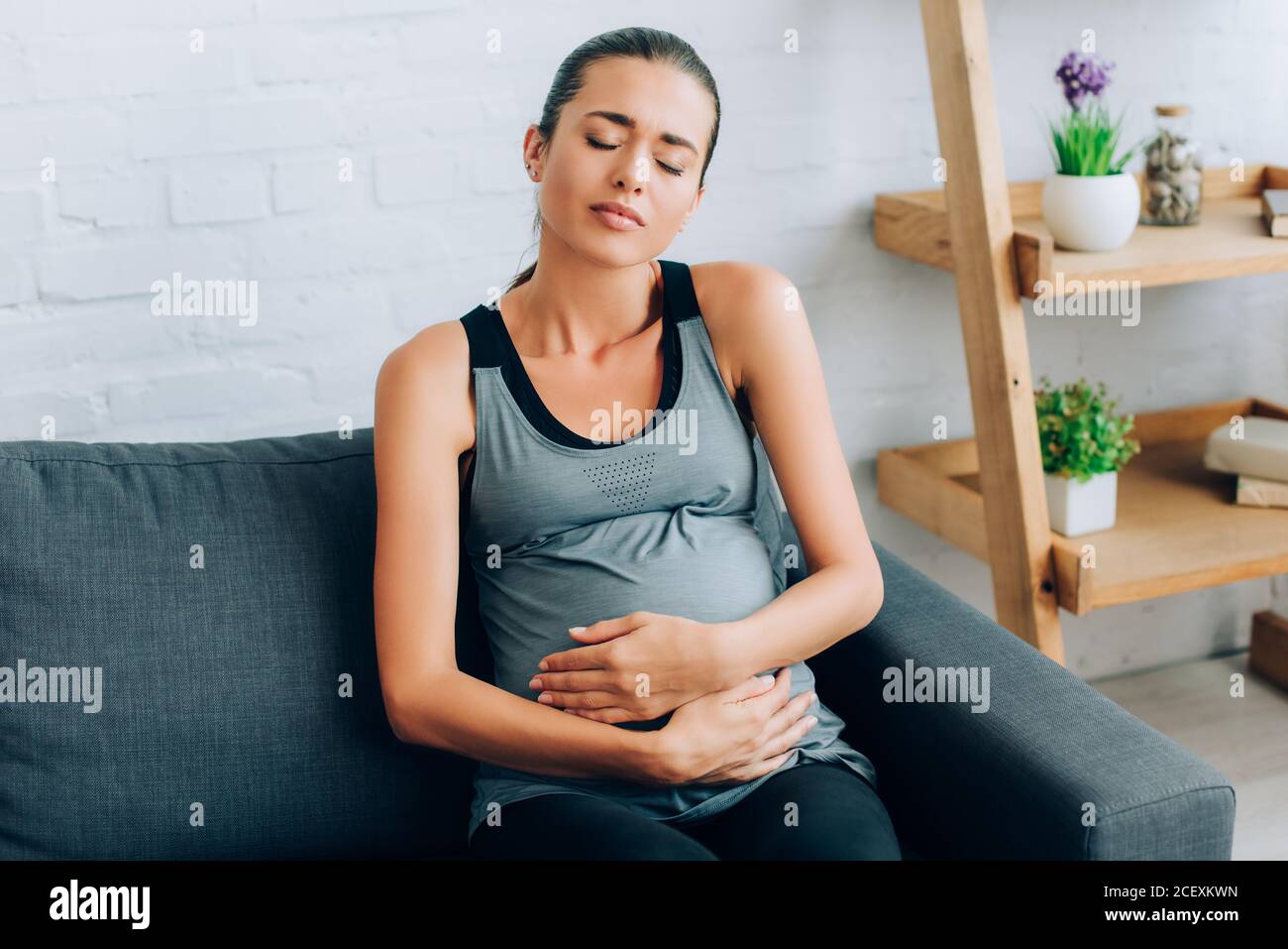 Tired sportswoman embracing belly while sitting on couch at home Stock ...