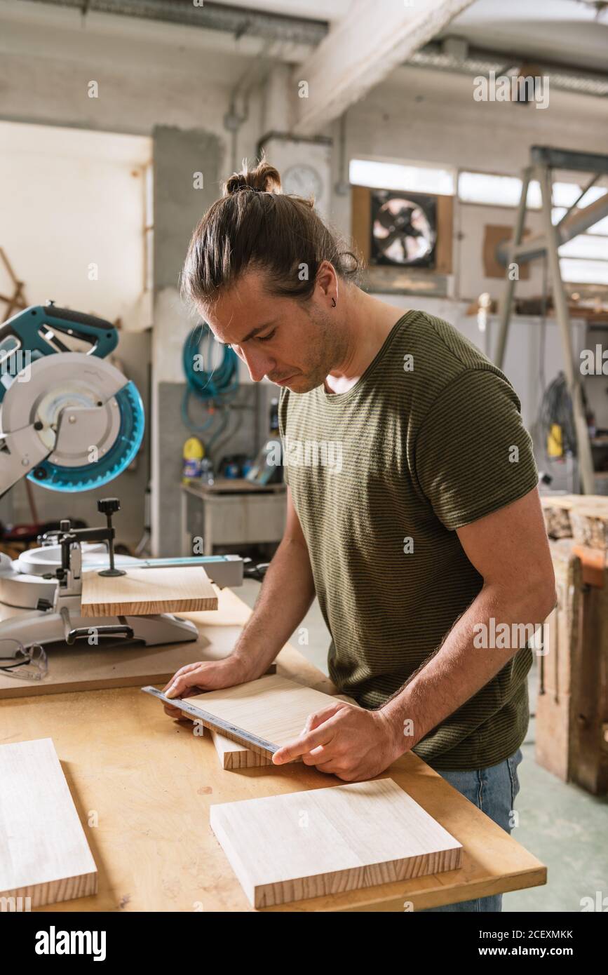 Male carpenter standing at workbench and measuring piece of wood with ...