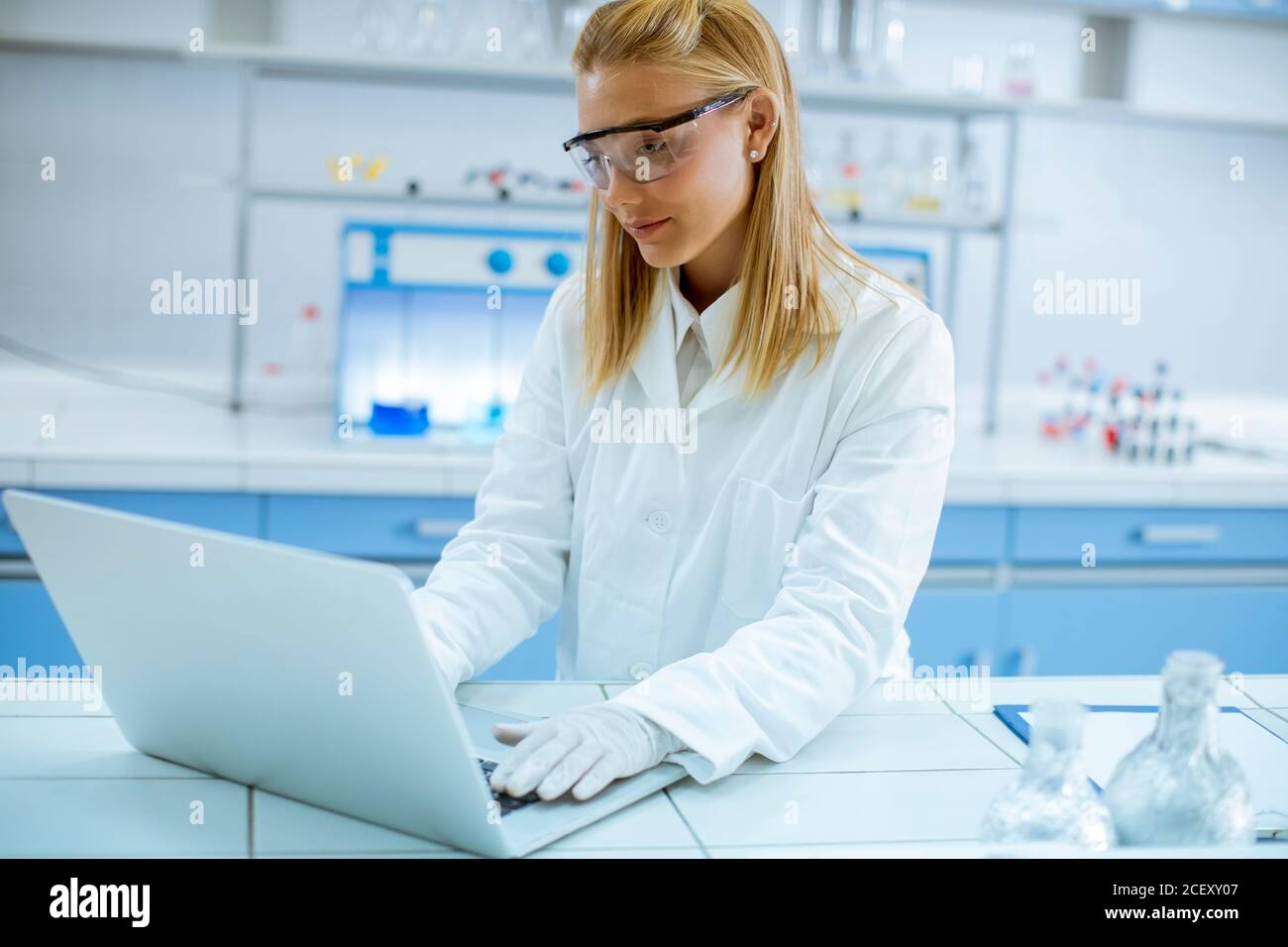 Cute female researcher in white lab coat and protective goggles using ...