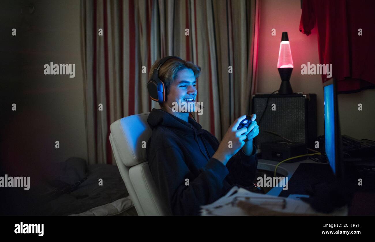 Smiling teenage boy playing video game at computer in dark bedroom ...