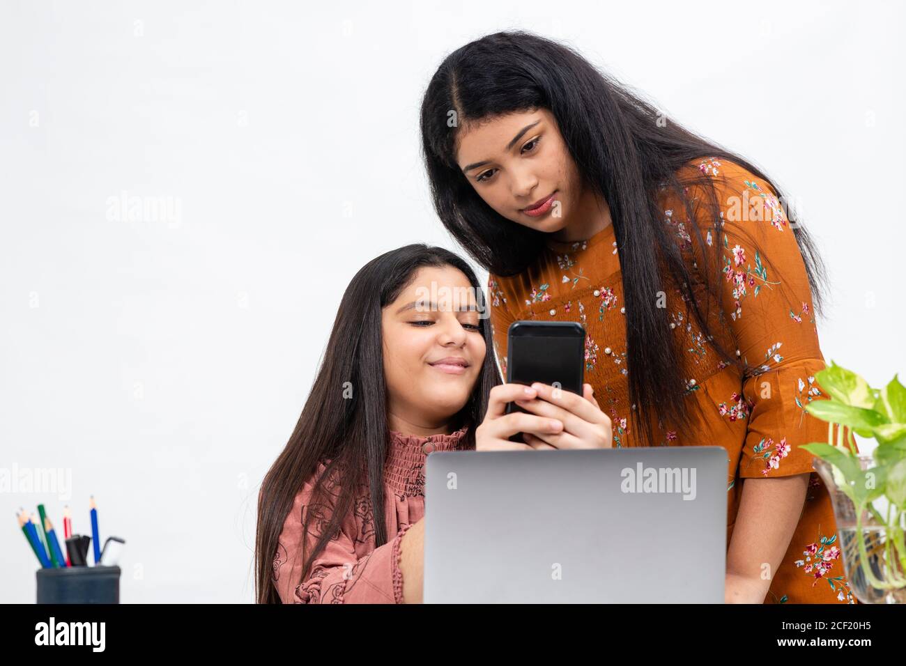 Two young Indian girls look into a phone with a laptop infront of them ...