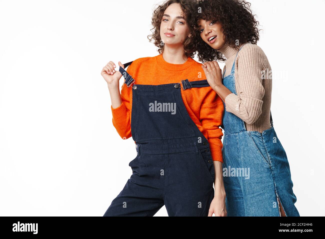 Portrait of two pleased women in overalls looking at camera and hugging ...