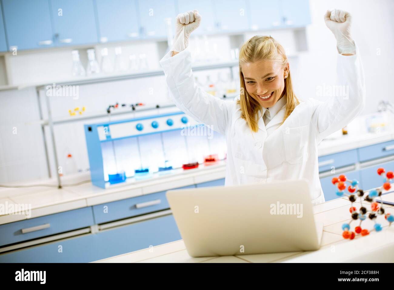 Cute happy female researcher in white lab coat using laptop while ...