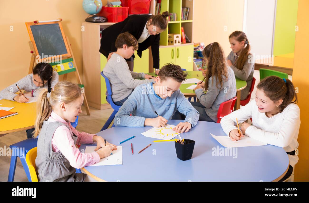 Smiling young boys and girls sitting and teacher helping drawing in ...