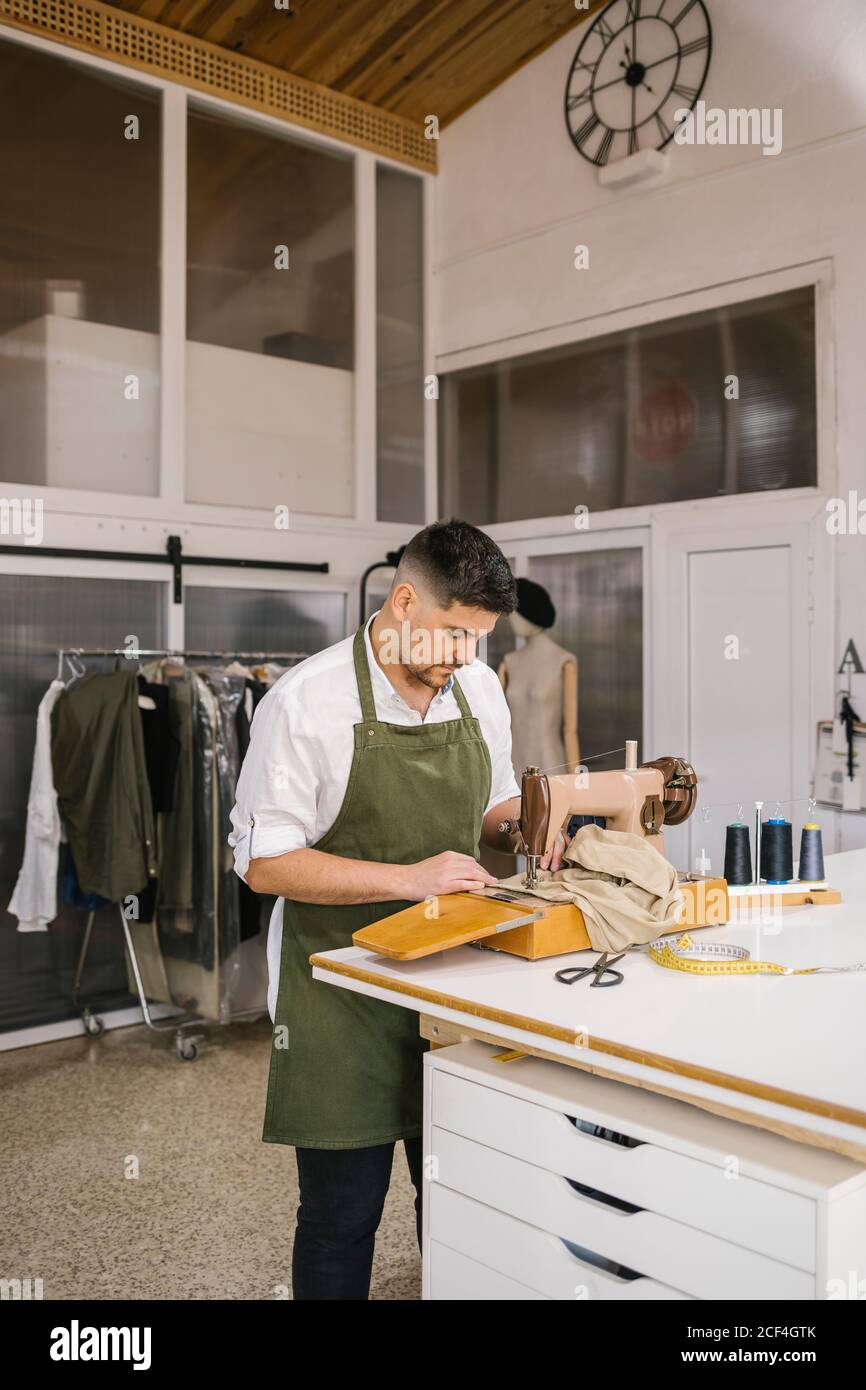 Diligent focused male tailor in apron sewing outfit details using ...