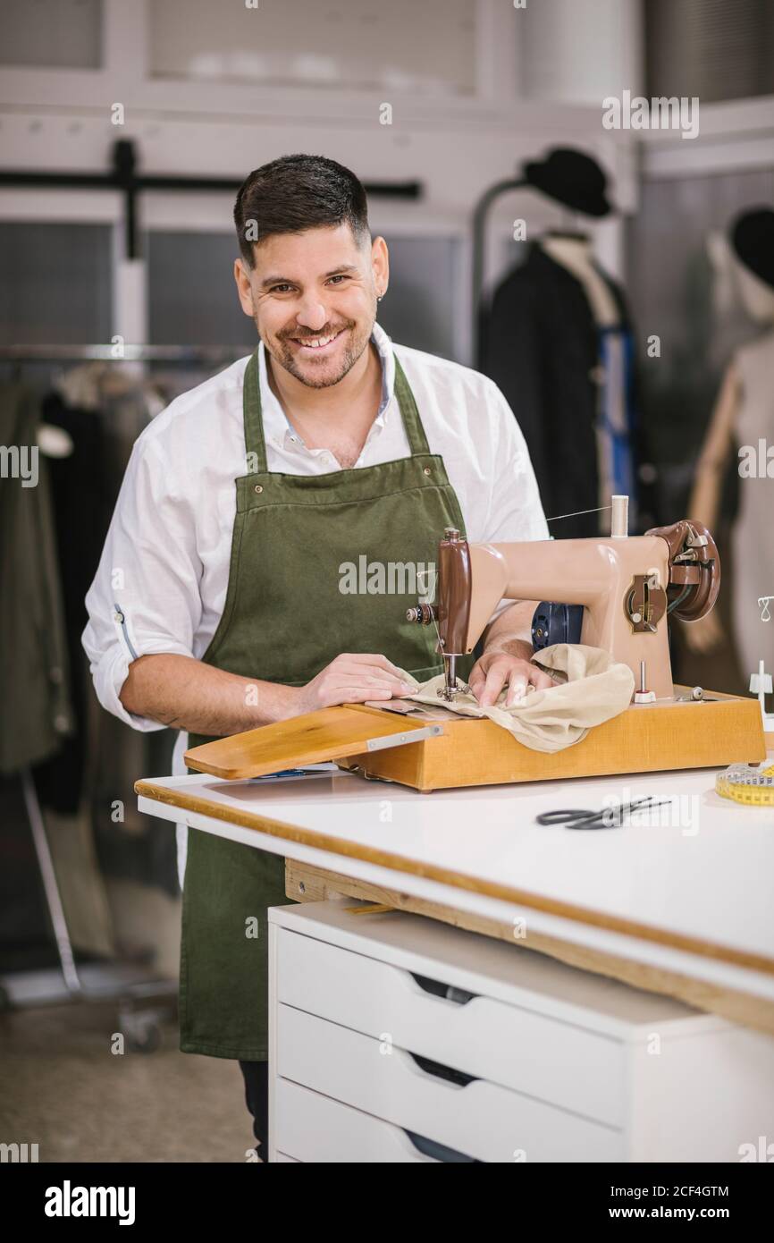 Diligent focused male tailor in apron sewing outfit details using ...