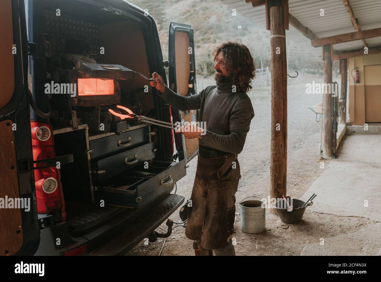 Bearded adult farrier taking hot horseshoe from portable furnace in ...
