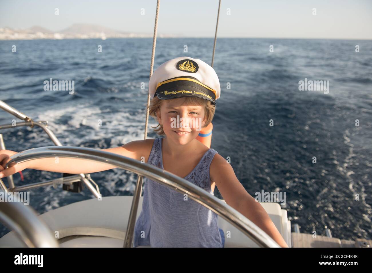 Positive kid in captain hat floating on expensive boat on sea in sunny ...