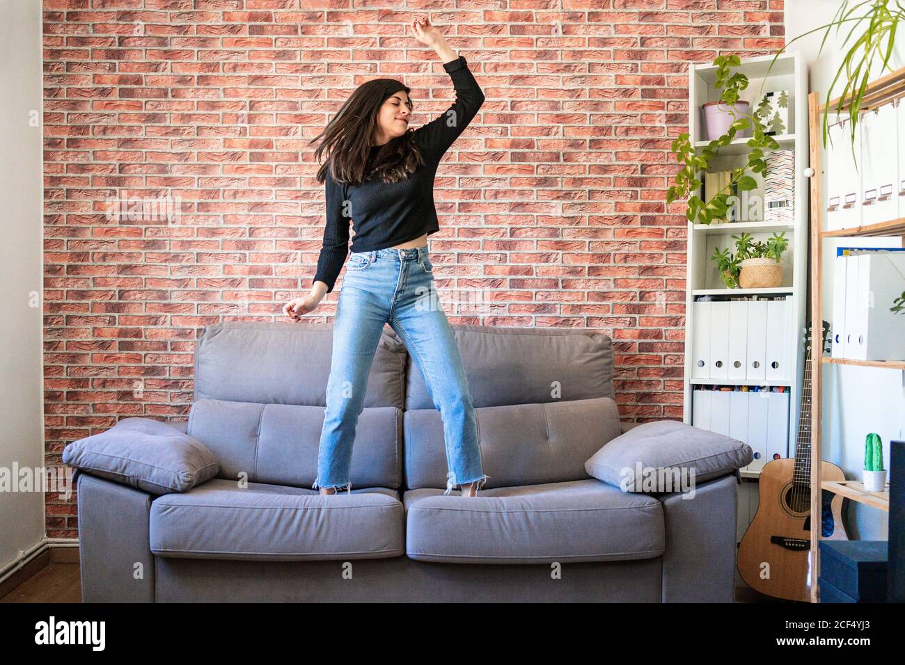 Young Woman dancing on a couch in her house. Behind it is a brick wall ...