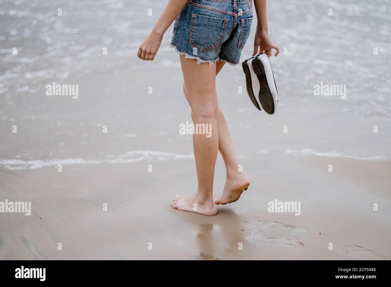 Side view of crop Woman in jean shorts walking with shoes in hand on ...