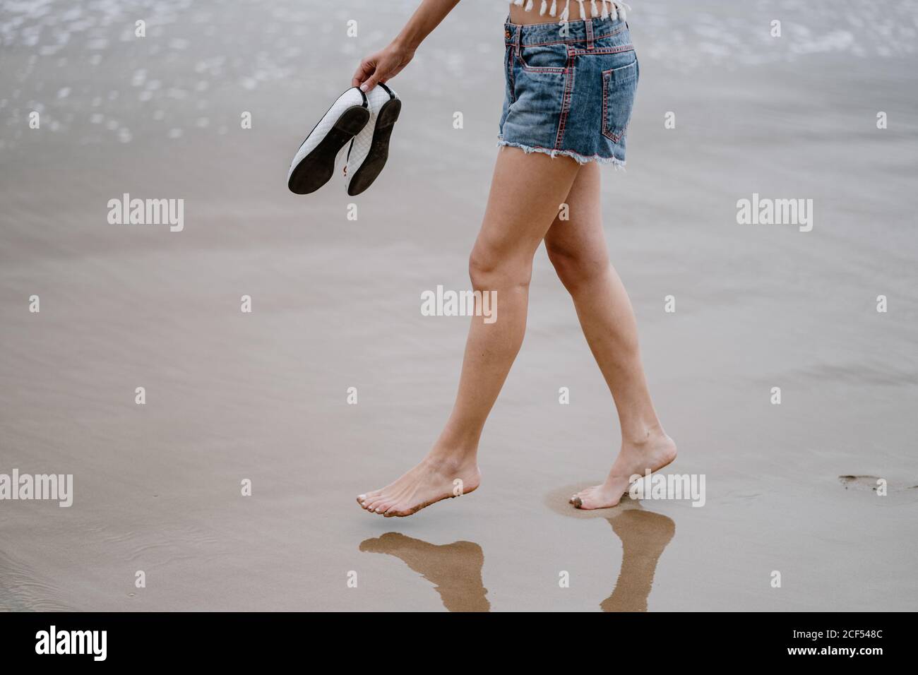 Side view of crop Woman in jean shorts walking with shoes in hand on ...