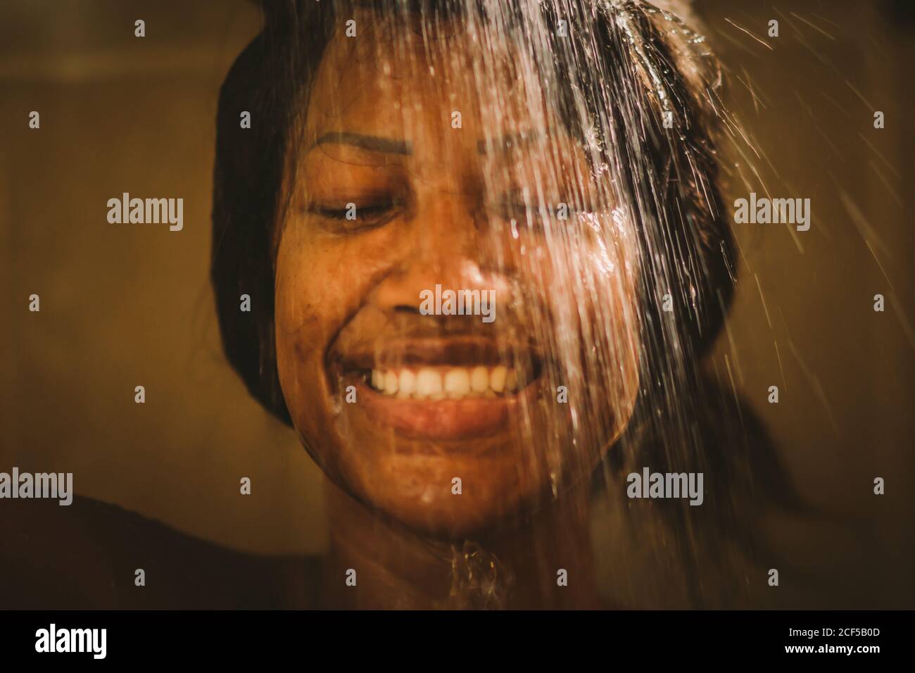 Beautiful ethnic Woman cheerfully smiling and keeping eyes closed while washing in shower at ...