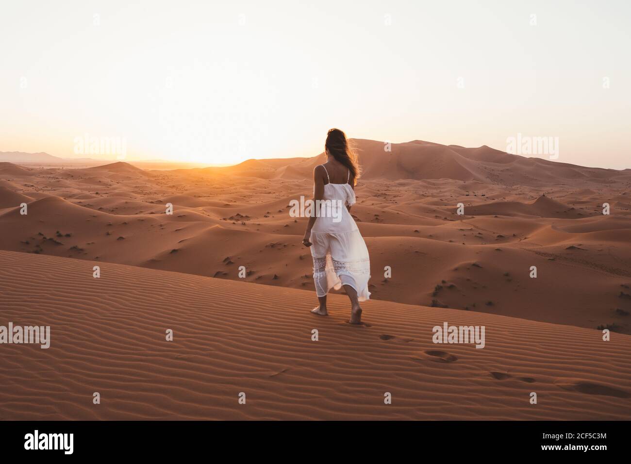 Back view of barefoot Woman in white summer dress walking on sandy dune of endless desert in ...