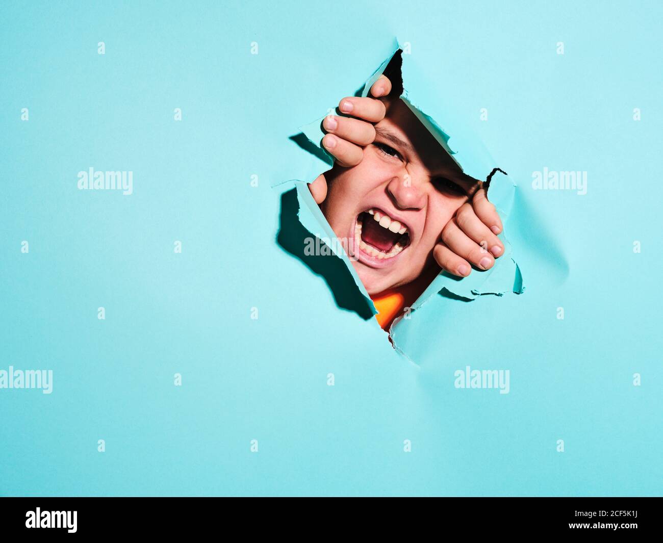 Screaming angry boy with grimace looking through ripped blue paper ...