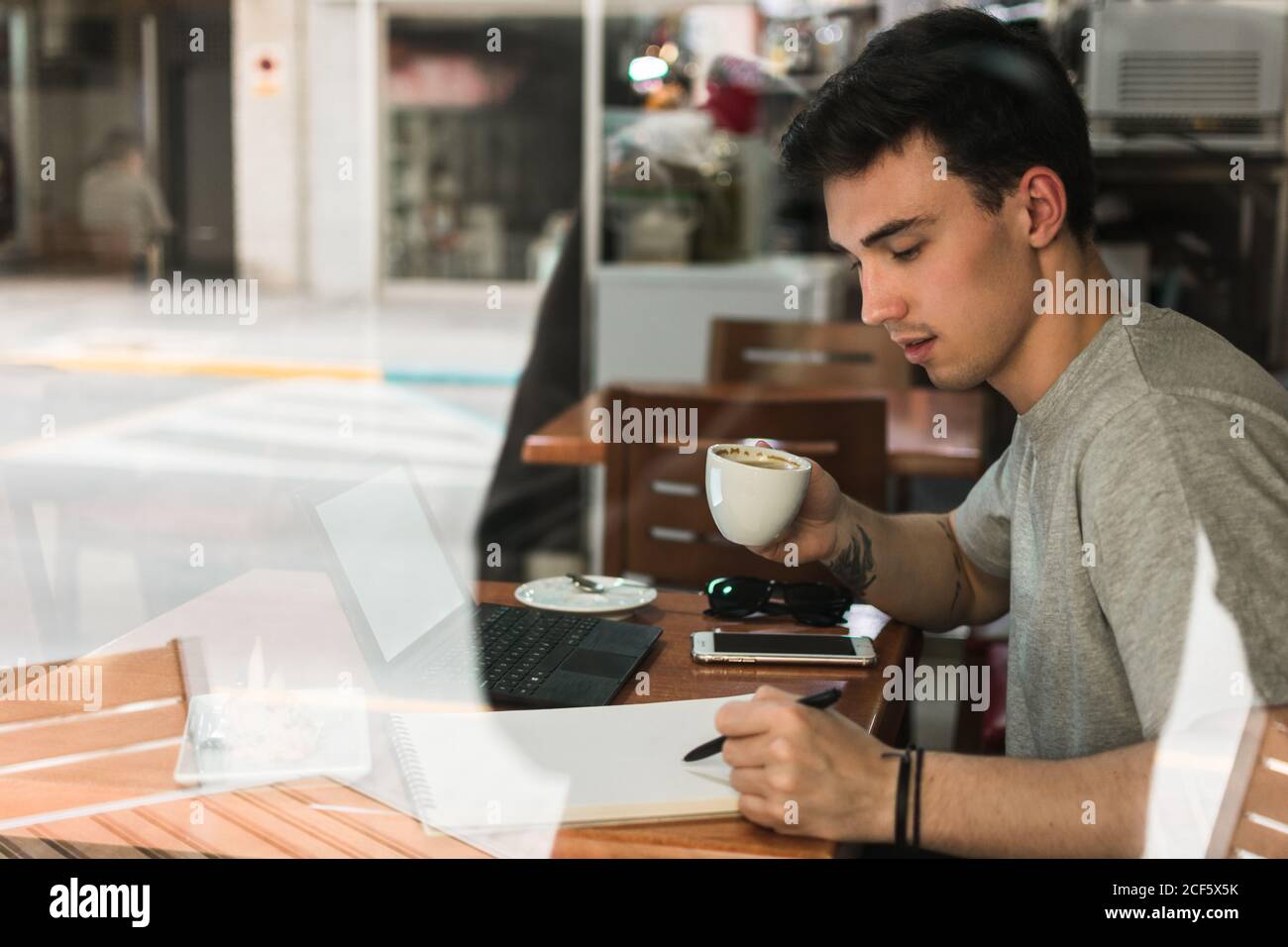 Side view of young man with cup of coffee writing in notepad while ...