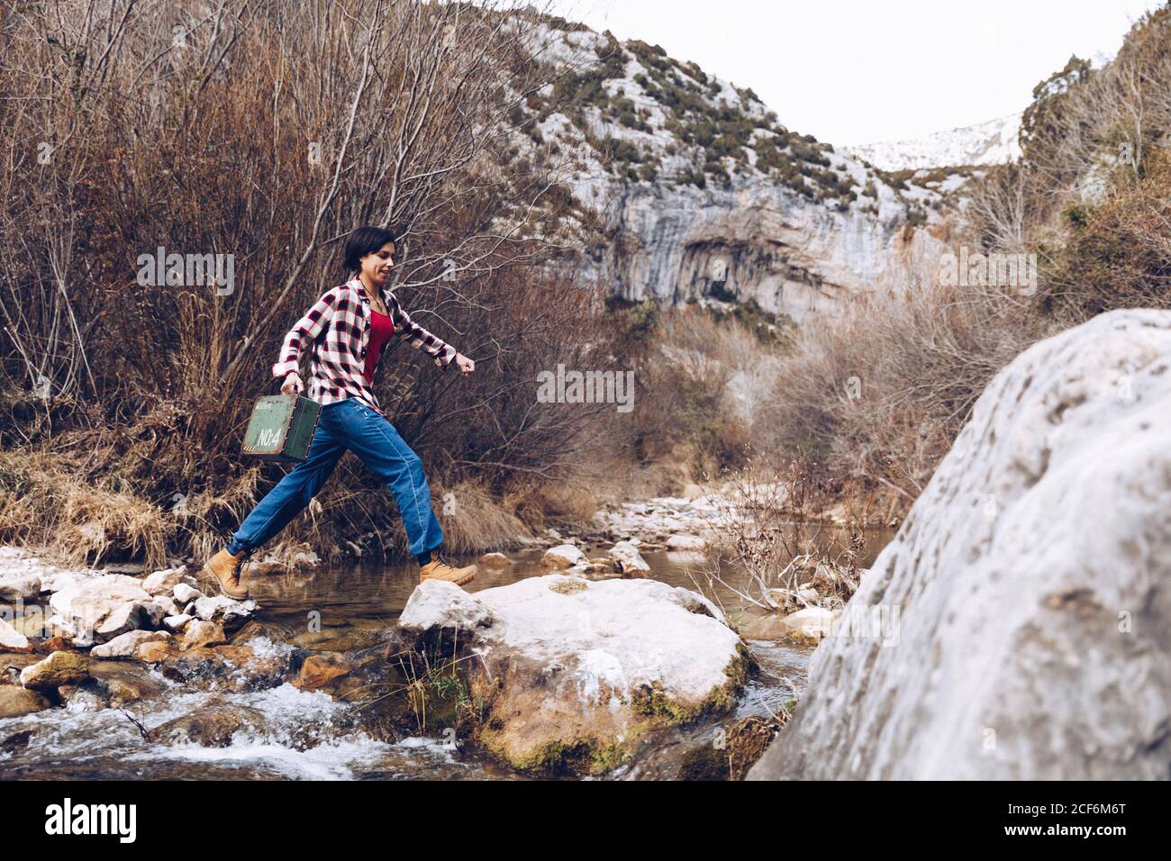 Side view of casual Woman carrying case and jumping on rocks of clear ...