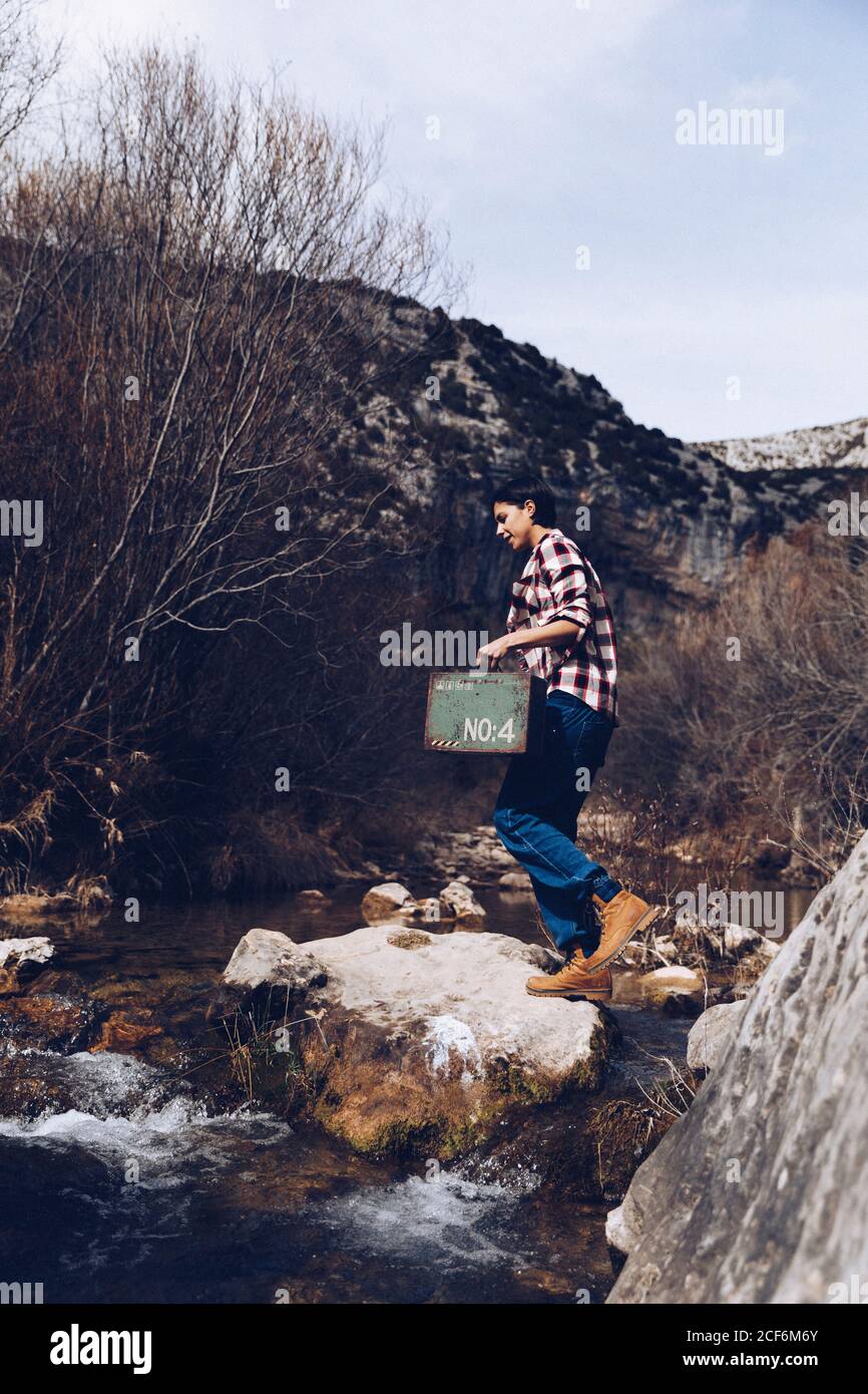 Side view of casual Woman carrying case and jumping on rocks of clear ...