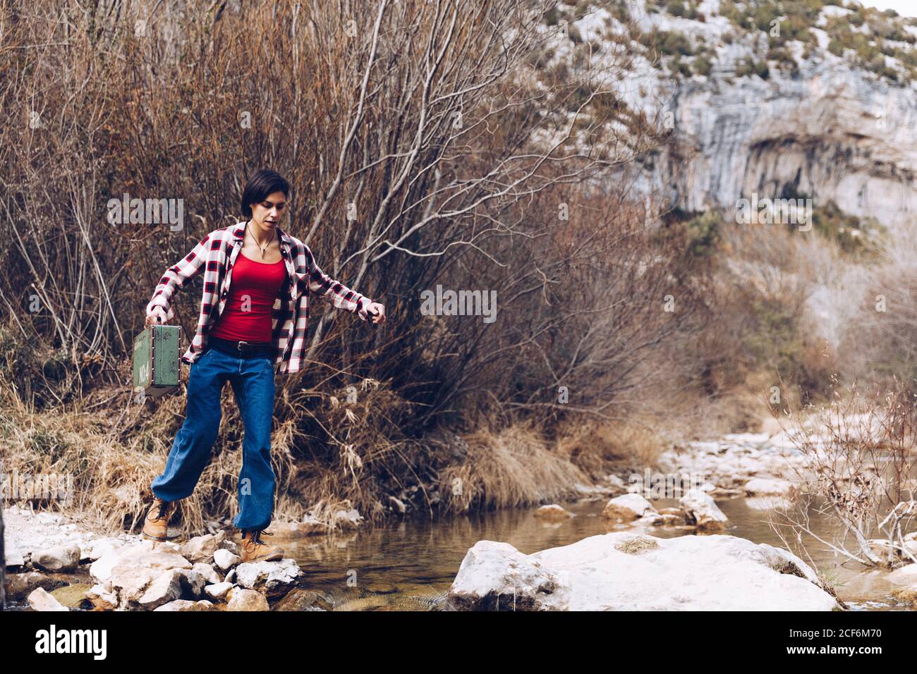 Side view of casual Woman carrying case and jumping on rocks of clear ...