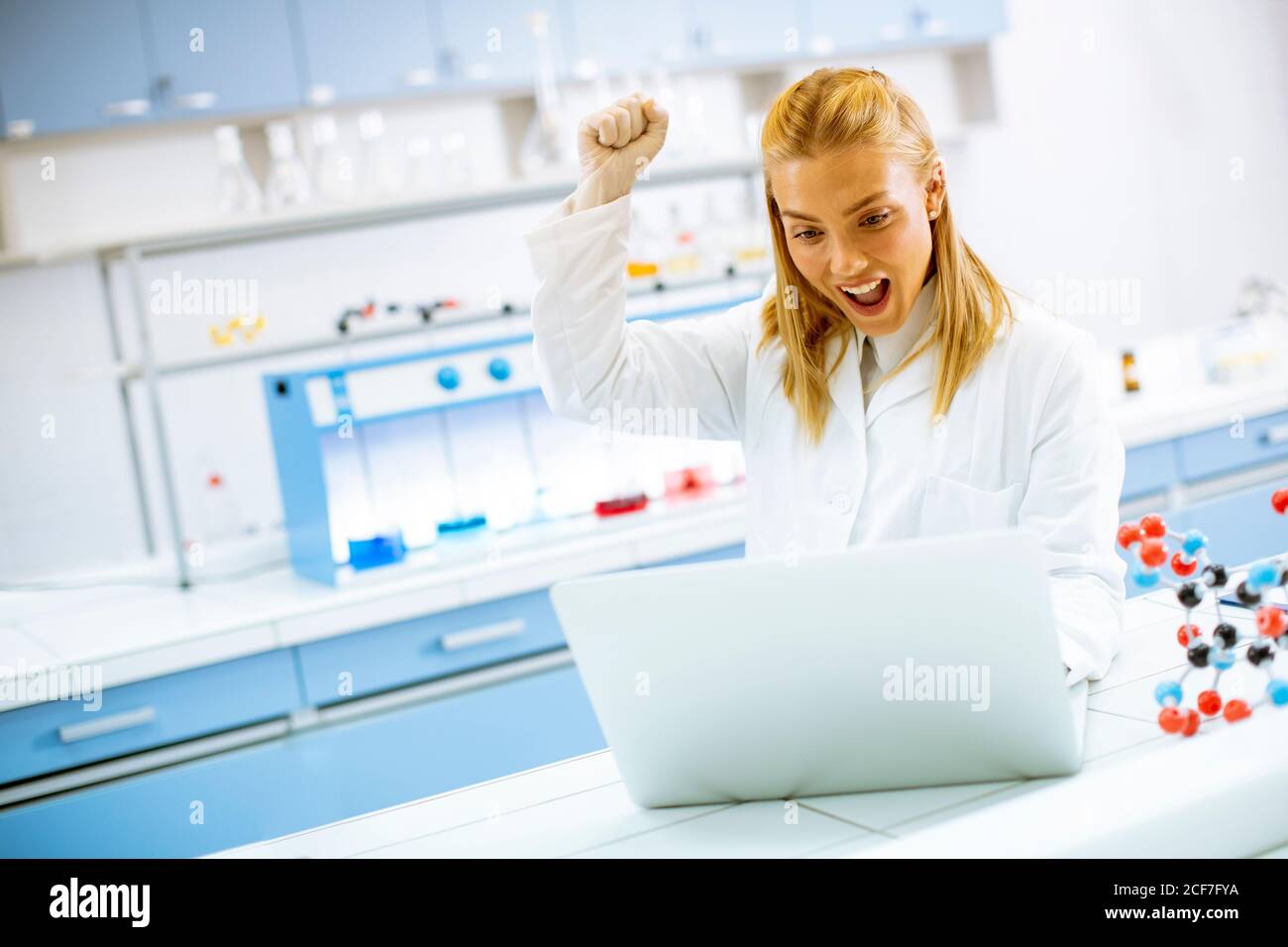 Cute happy female researcher in white lab coat using laptop while ...