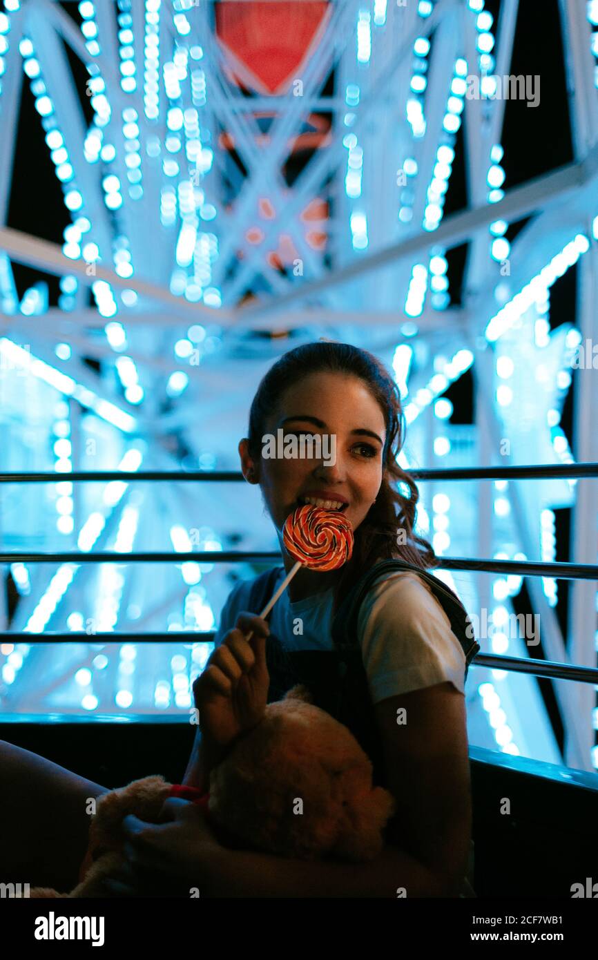 Pretty female teenager with teddy bear having fun in funfair and eating ...