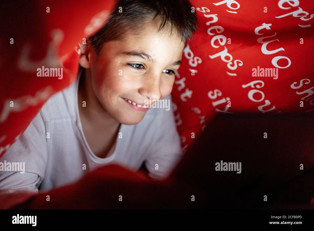 Adorable kid in pajamas hiding under blanket and using tablet while ...