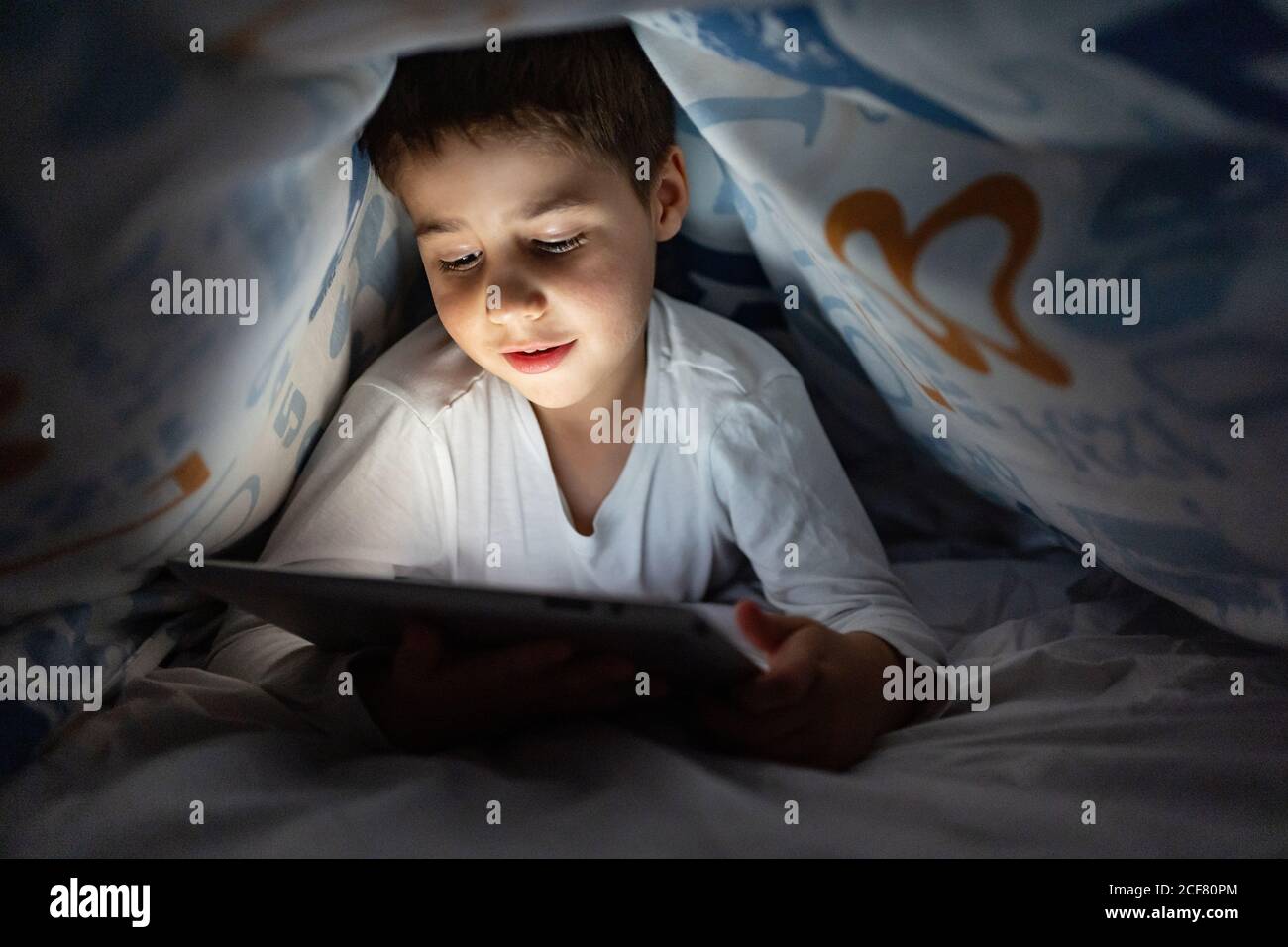 Adorable kid in pajamas hiding under blanket and using tablet while ...