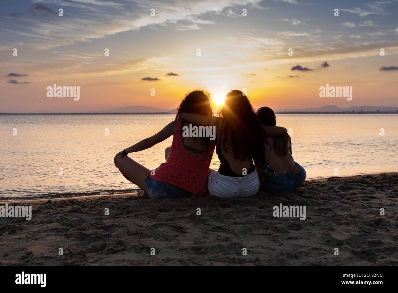 Back view of bronzed female friends hugging and relaxing on beach at ...