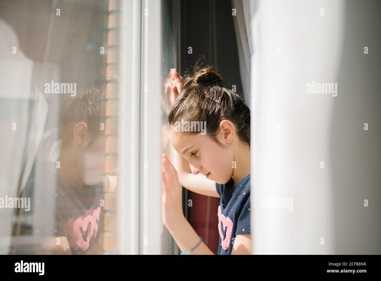 Side view of sad girl standing near window with hands on glass and ...