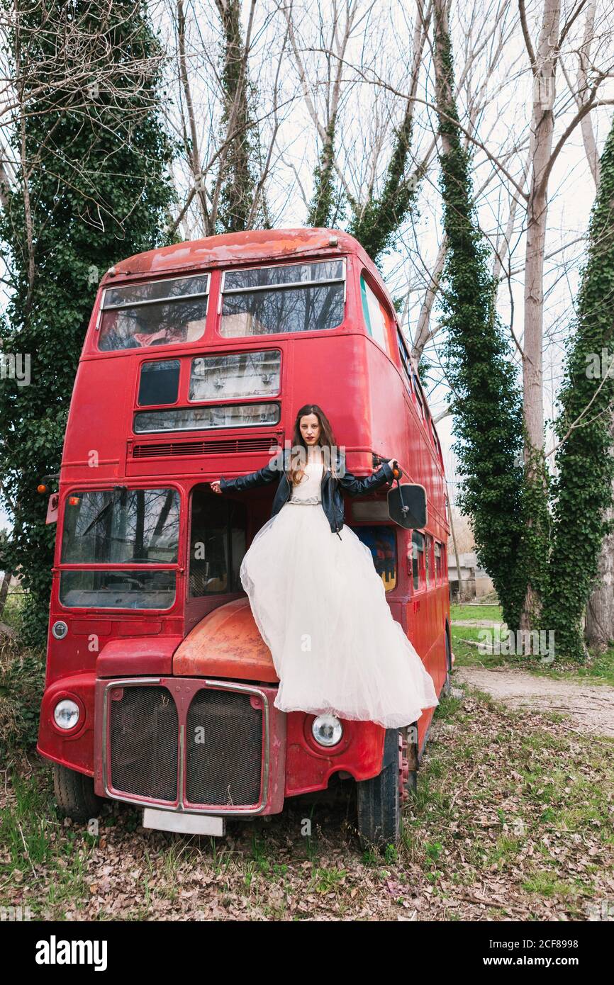 Elegant newlywed female wearing white dress sitting on hood of retro ...