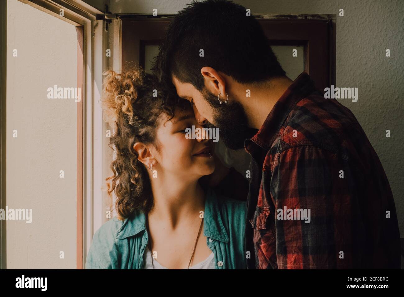 Joyful casual couple smiling and hugging while leaning on window sill ...