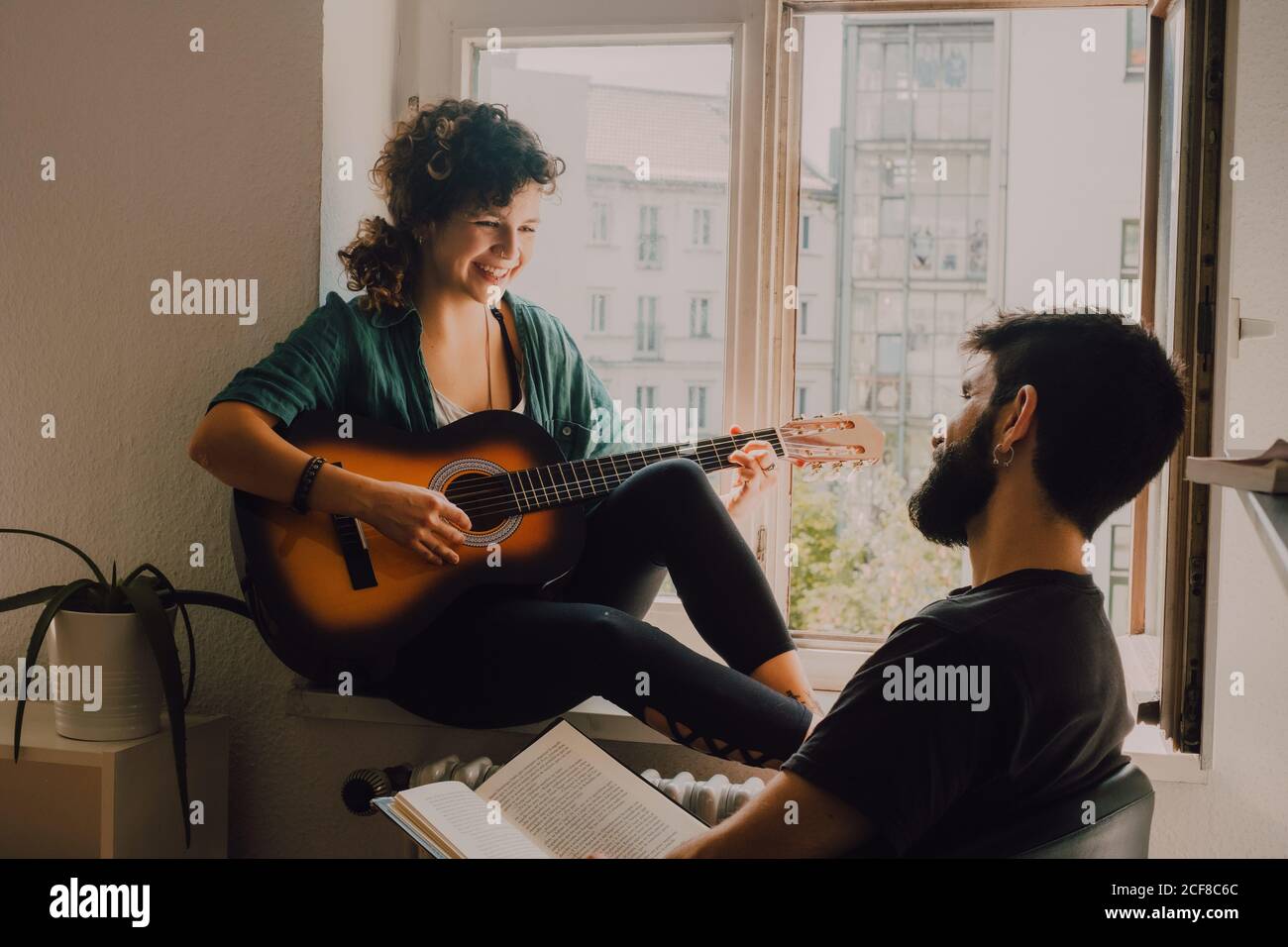 Happy Woman playing guitar and sitting on window sill while man reading ...