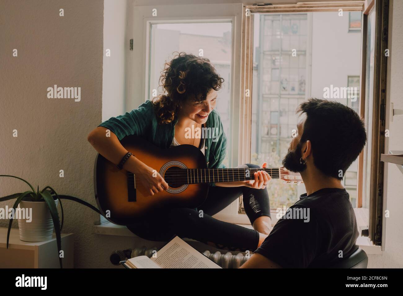 Happy Woman playing guitar and sitting on window sill while man reading ...