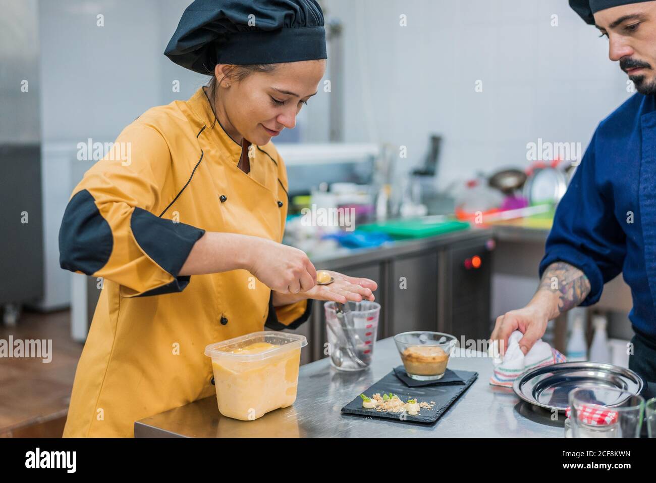 Male chef watching young female assistant with spoon in hand putting ...
