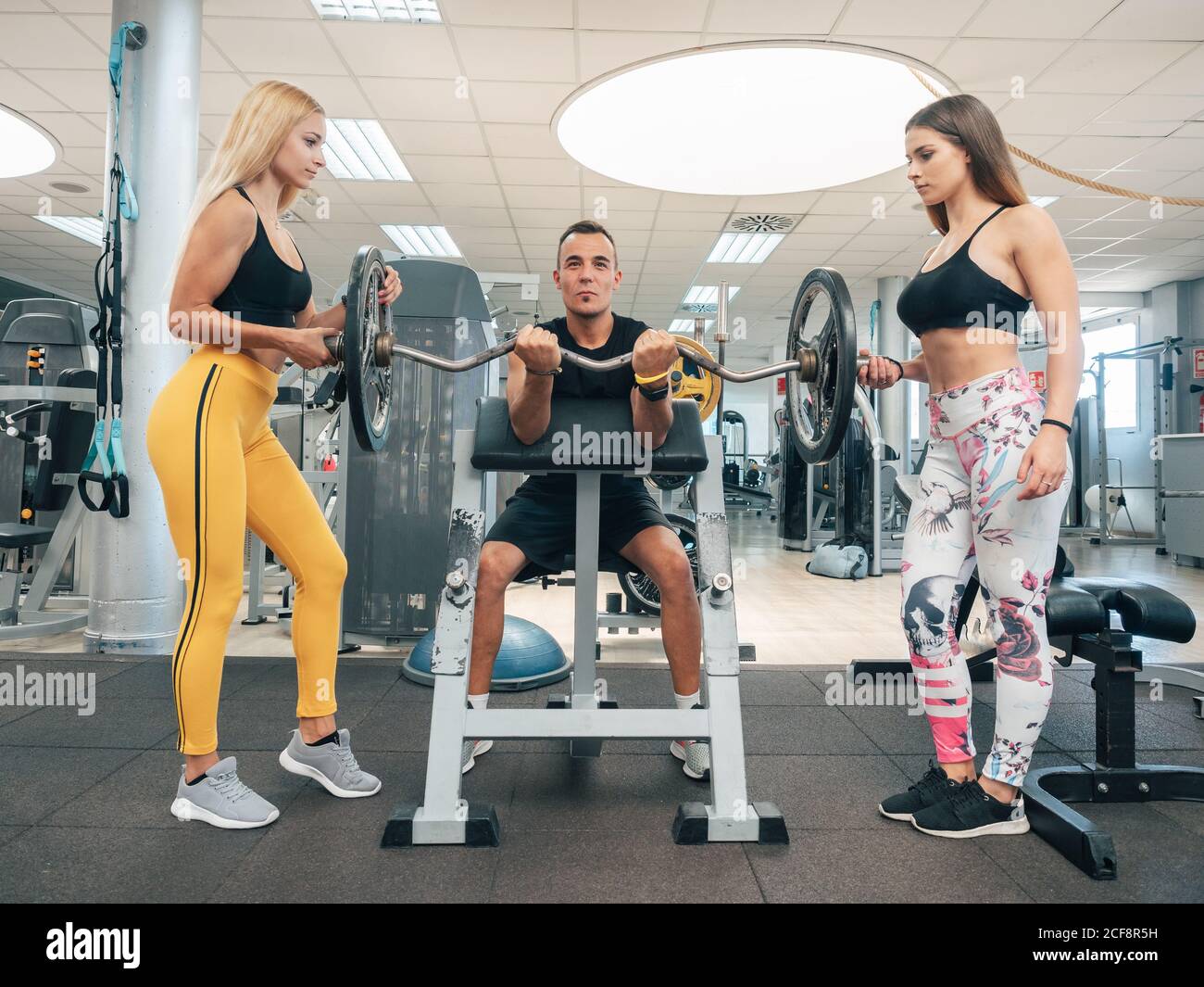 Strong male doing bicep curls on bench with young slim women holding ...