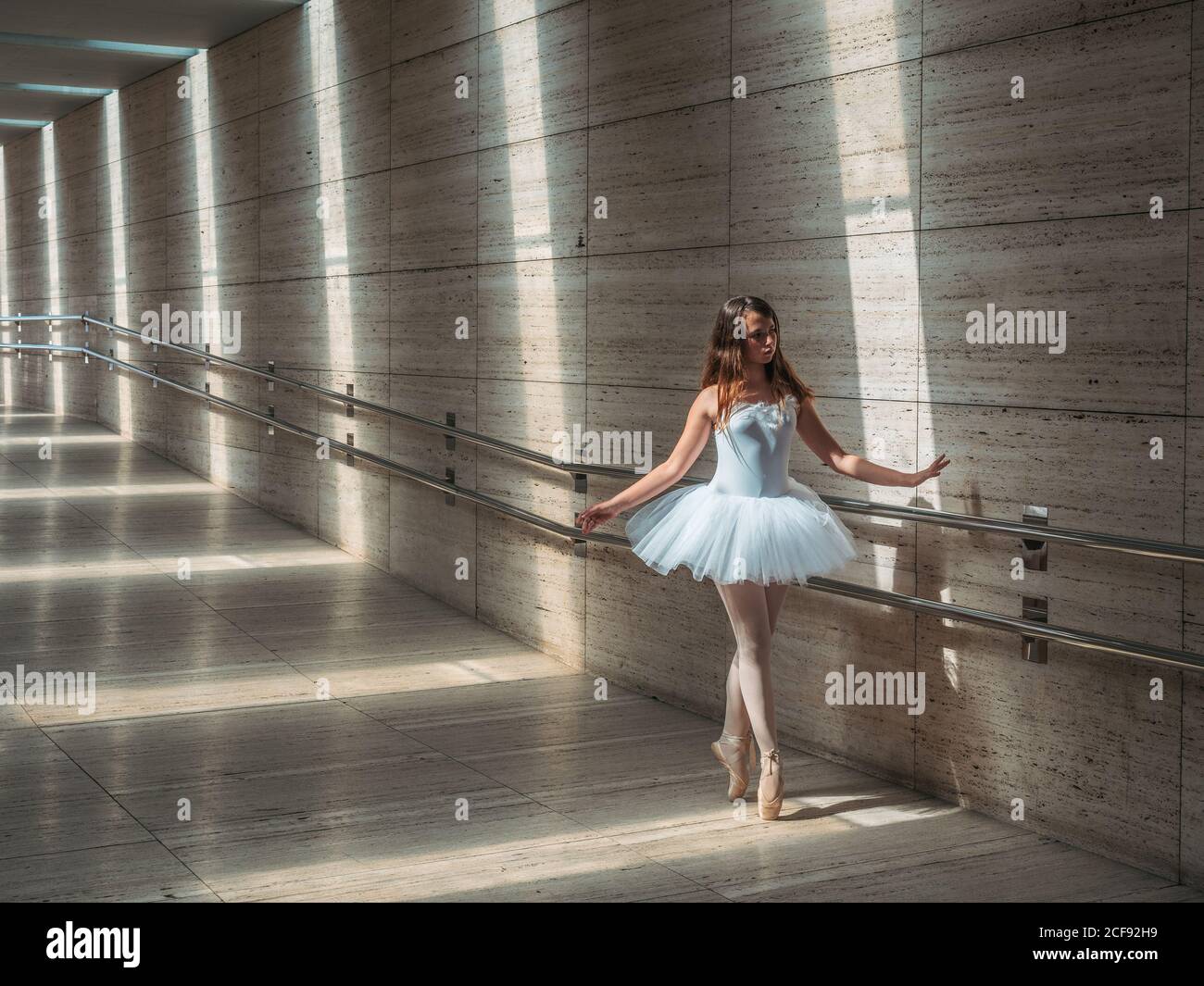 Ballerina in white ballet tutu performing exercises at rail in contrast ...